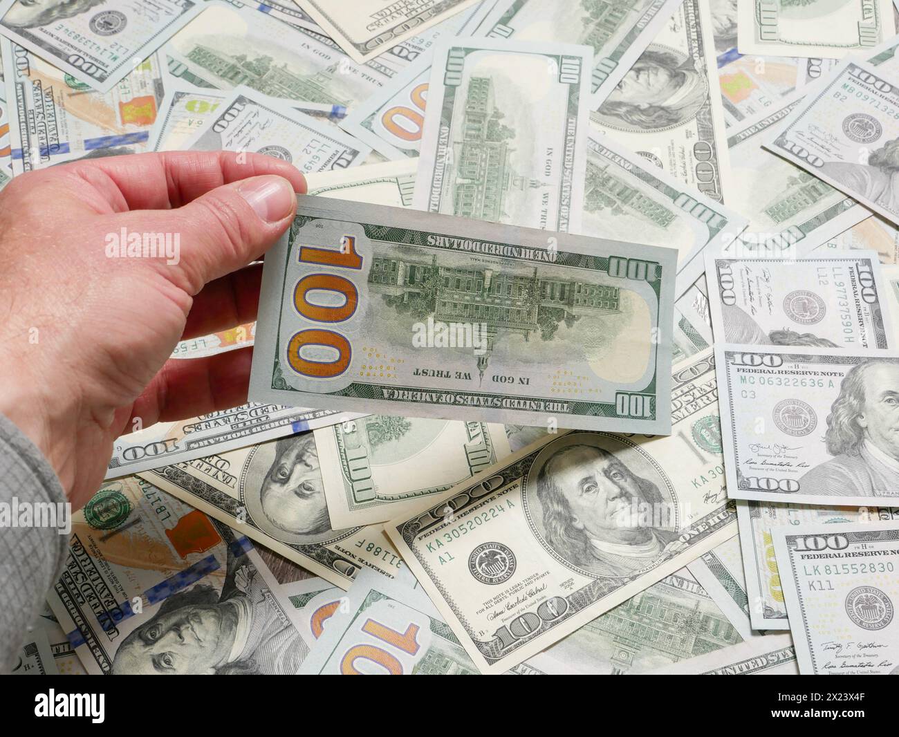 A man's hand holds one 100 dollar bill against the background of a pile ...