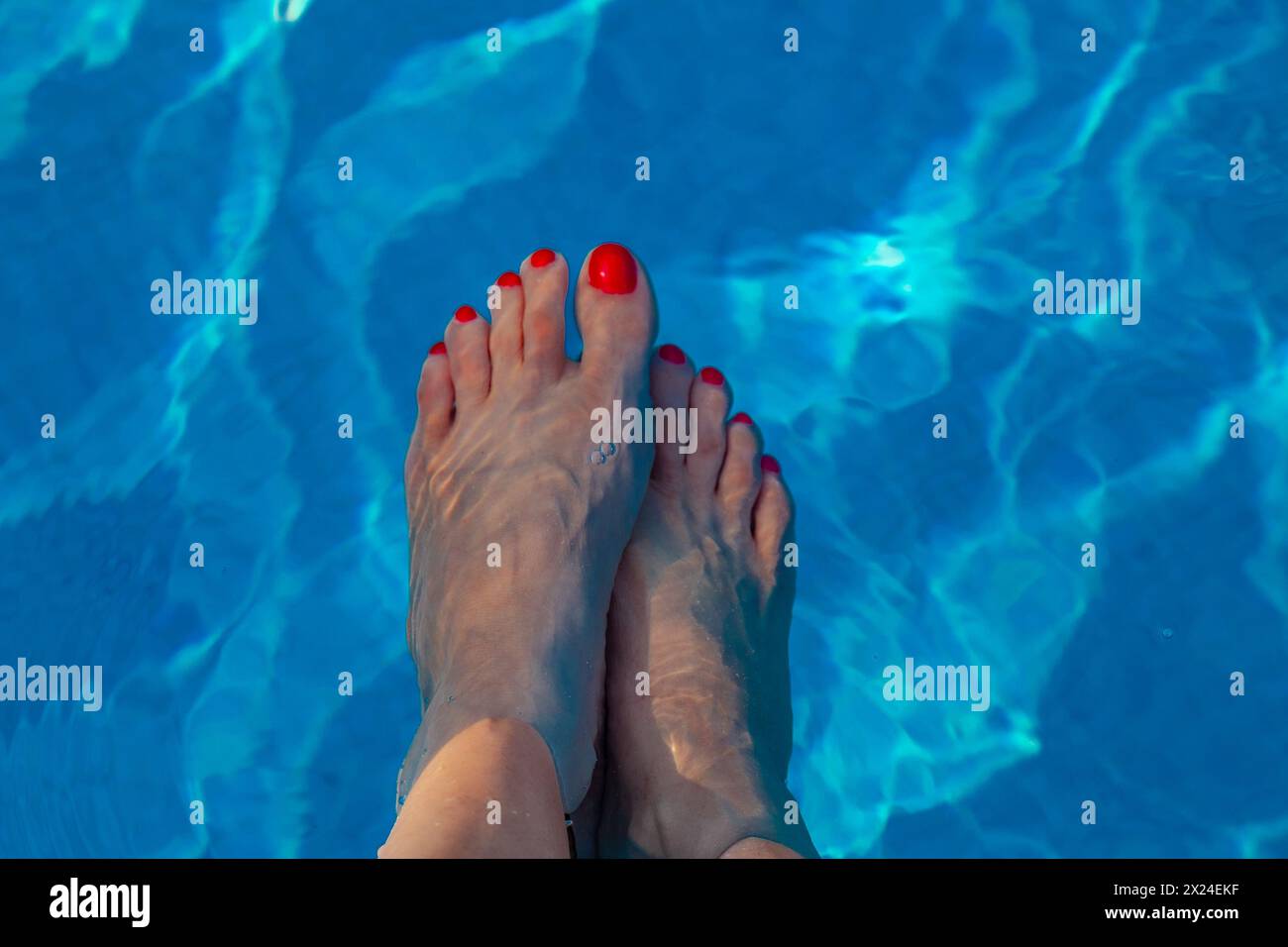 Female feet with red polished toe nails immersed in a blue water pool ...