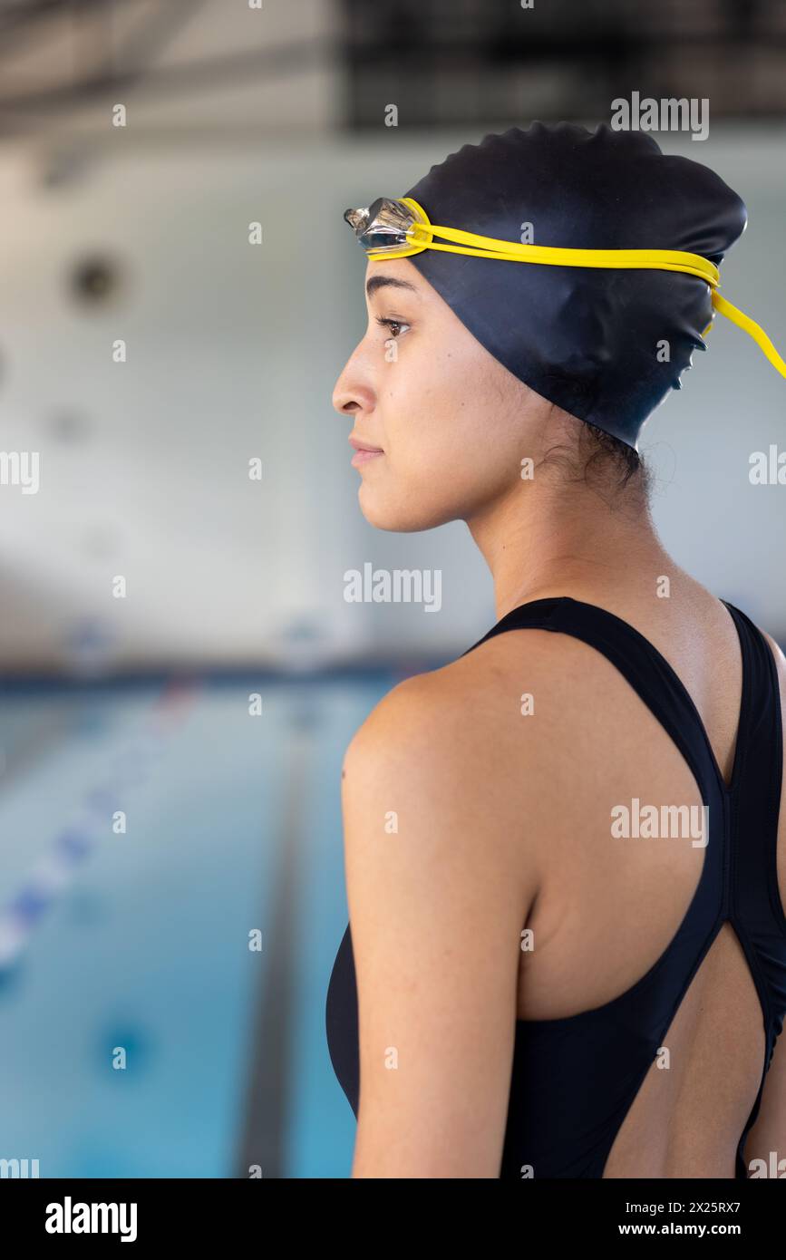 Biracial young female swimmer standing by poolside indoors, looking ...