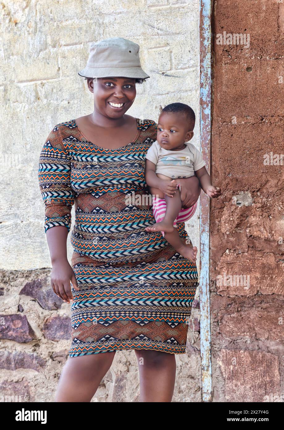 young village mother holding her baby, wearing a hat, standing in front ...