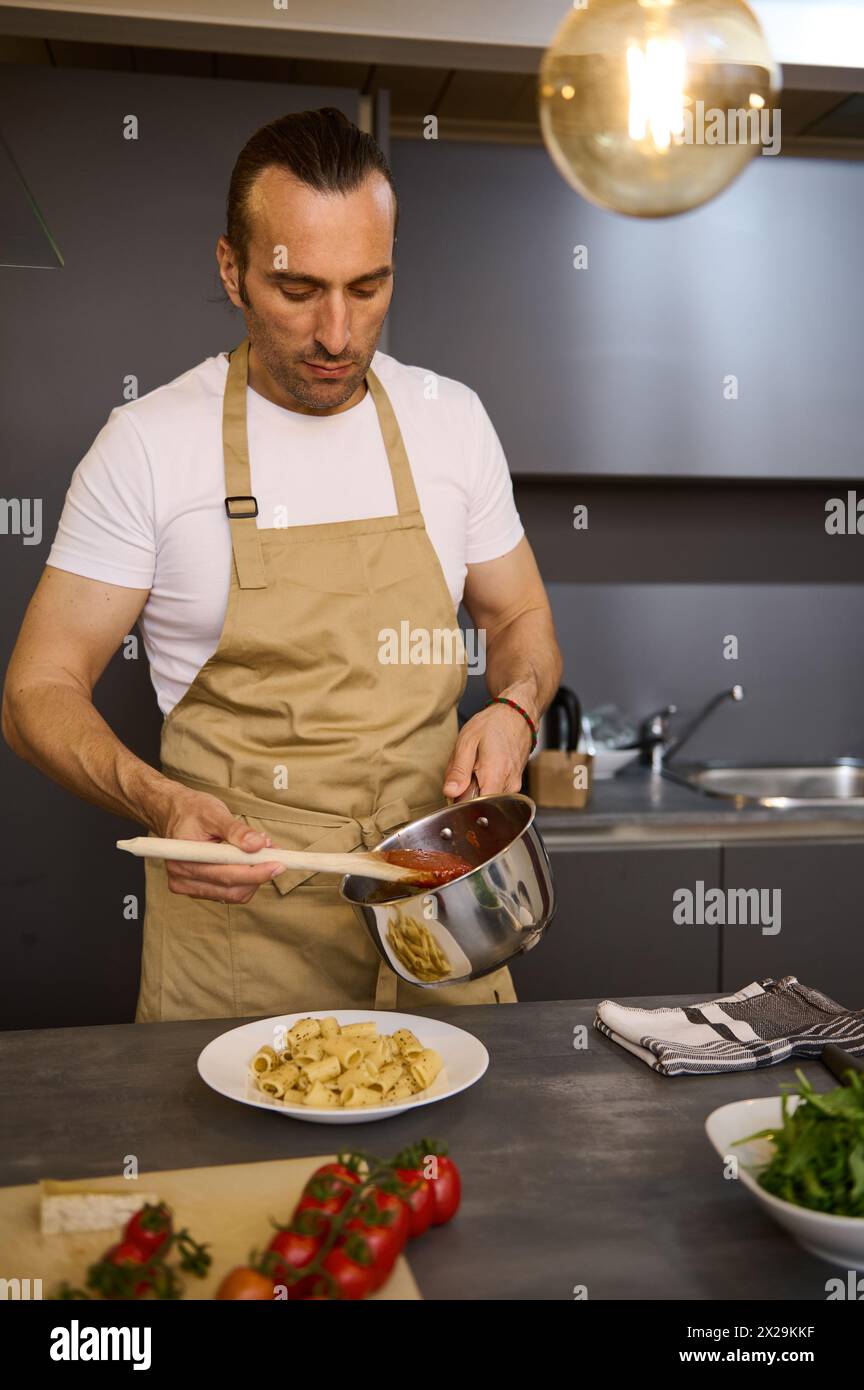 Vertical shot of a handsome European man in chef apron, pouring tomato ...