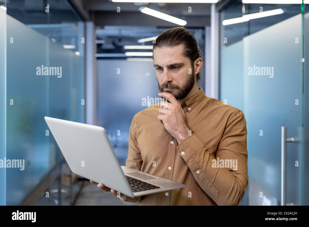 A thoughtful male programmer holding a laptop and analyzing data in a contemporary office ...