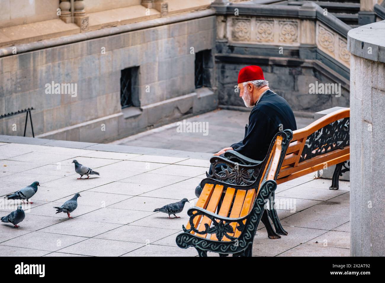 Old orthodox monk sitting on a bench, wearing a red skufia and feedeing ...
