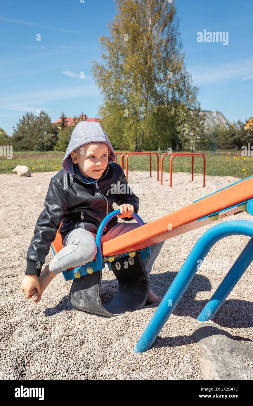 A barefoot little boy sits on an orange balance swing in an outdoor ...