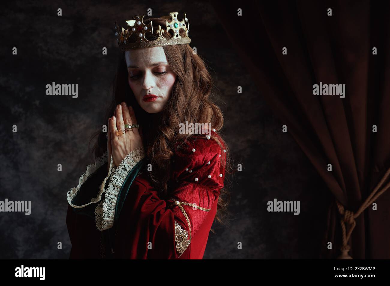medieval queen in red dress with crown praying on dark gray background ...