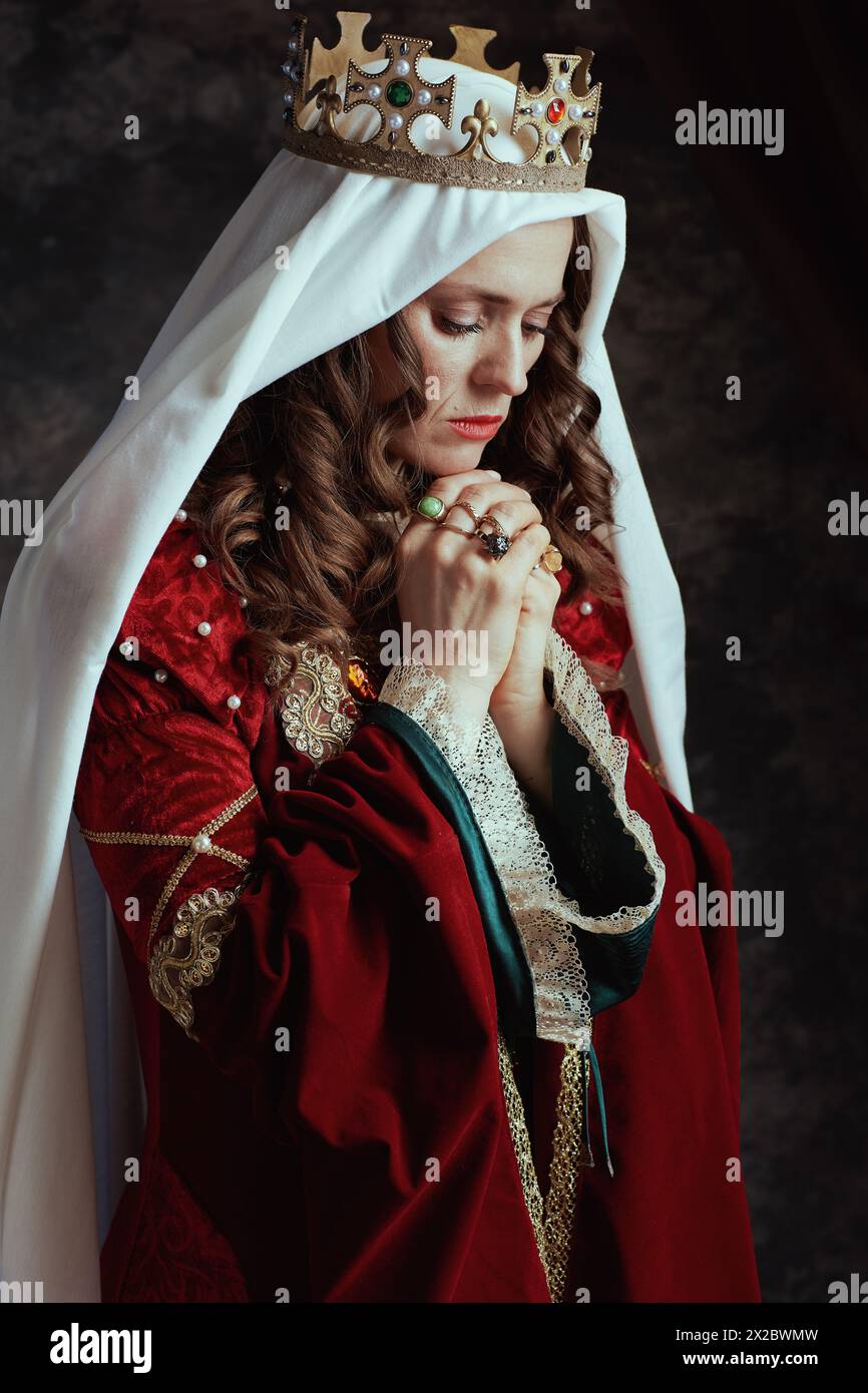 medieval queen in red dress with veil and crown praying on dark gray ...