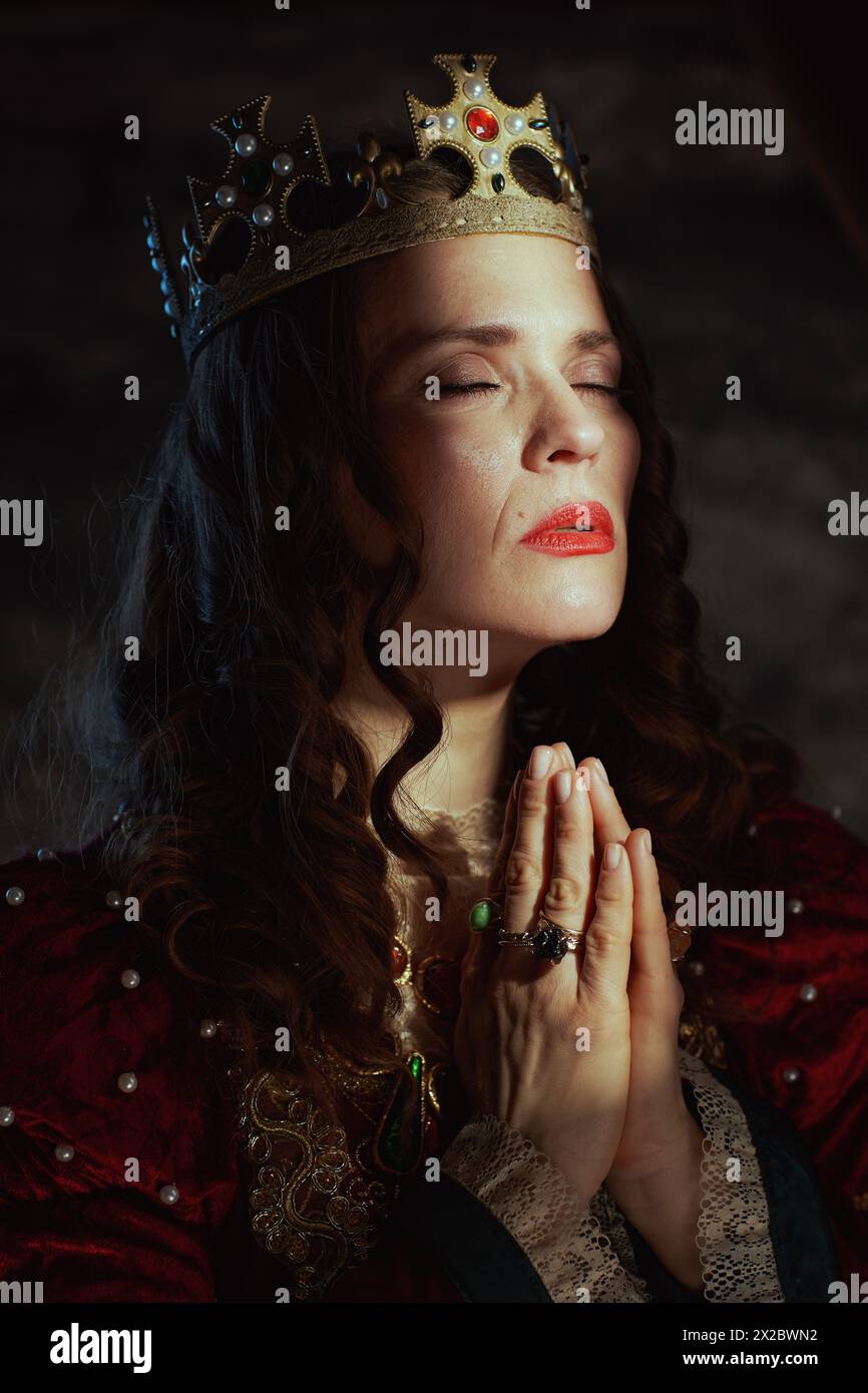 medieval queen in red dress with crown praying on dark gray background ...
