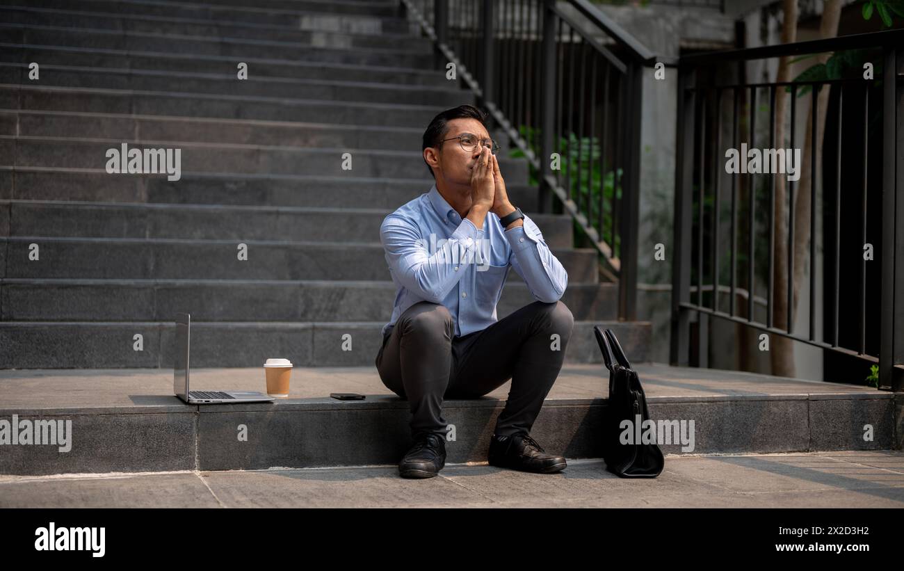 An Asian millennial businessman sits on outdoor steps with a laptop, a ...
