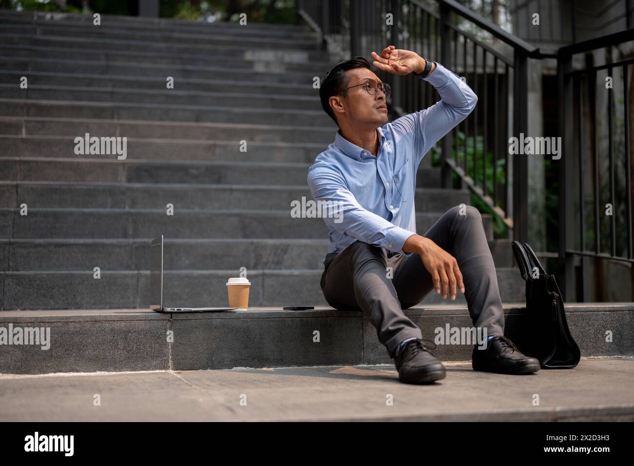 An Asian businessman sits on outdoor steps, looking upward with his ...