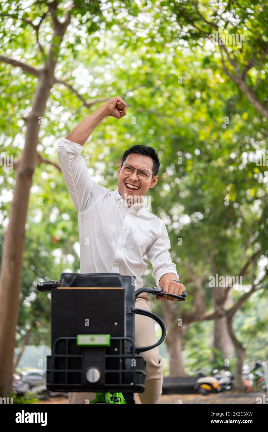 An excited Asian millennial businessman rides a bike triumphantly ...