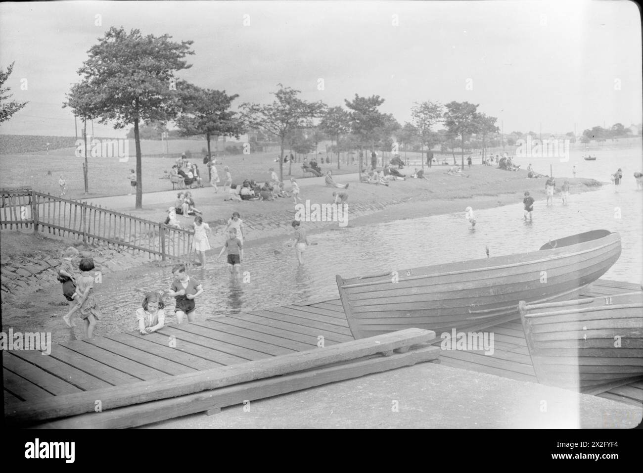 Children paddle rowing boats and sit along the shaded, tree-lined bank ...