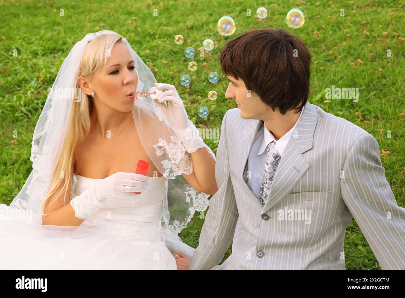 Beautiful young bride wearing white dress sitting on green grass with groom and blowing bubbles ...