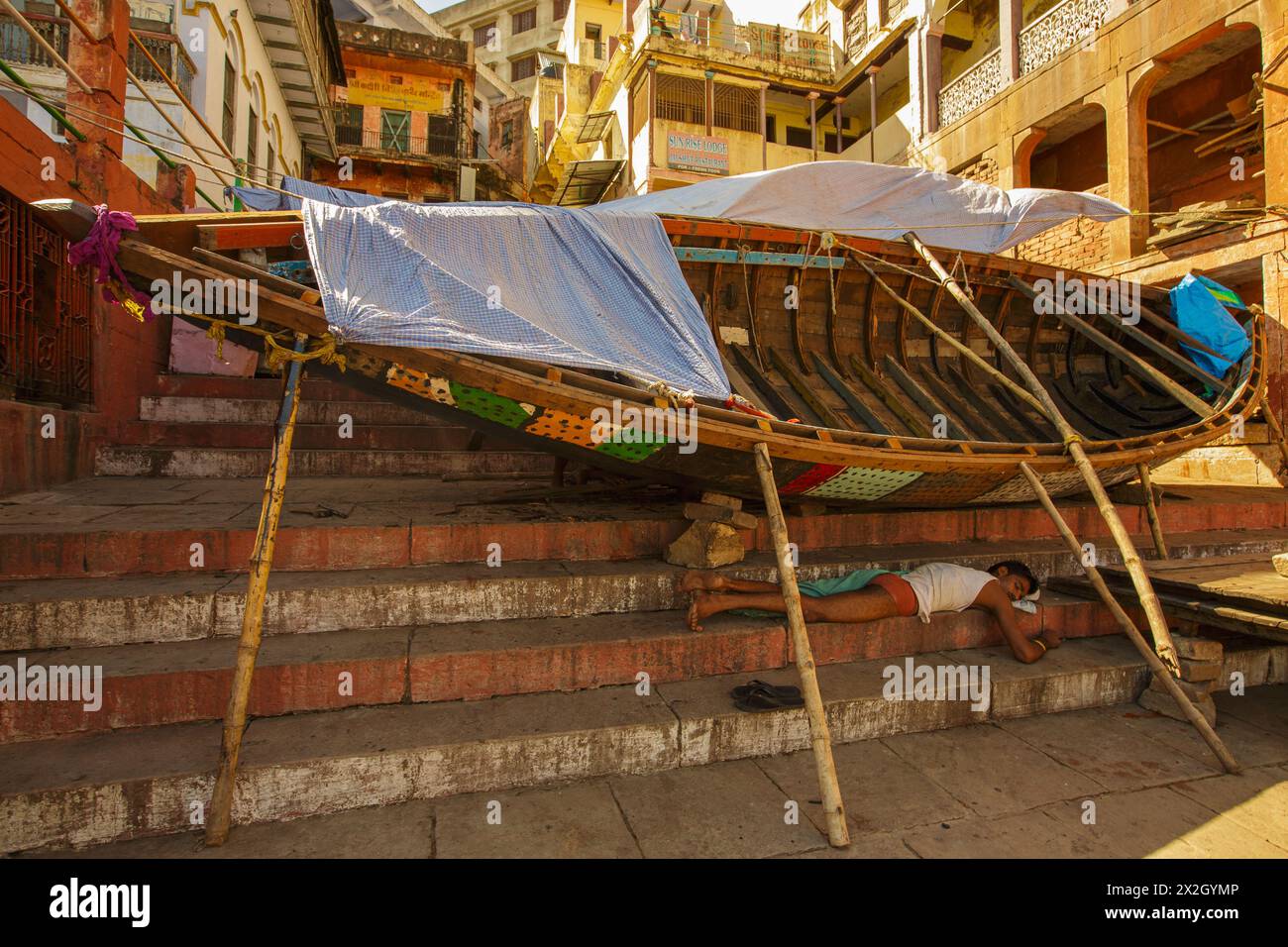 Workman sleeping under a boat pulled up onto Meer Ghat for repairs in ...