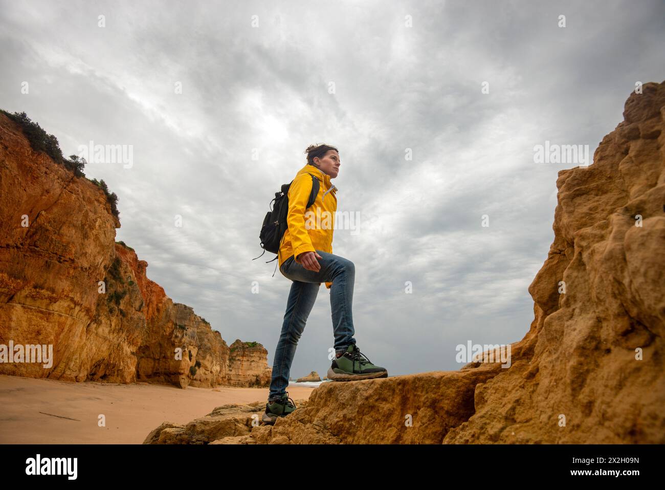 Woman hiker with backpack walking up rocks by a beach, wearing a yellow ...