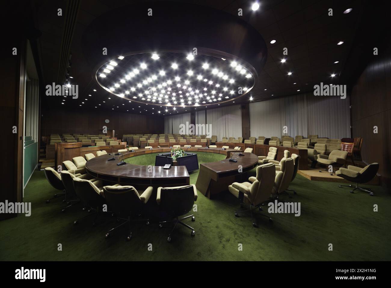 Auditorium with round oak table and beige armchairs, rows of desks and ...