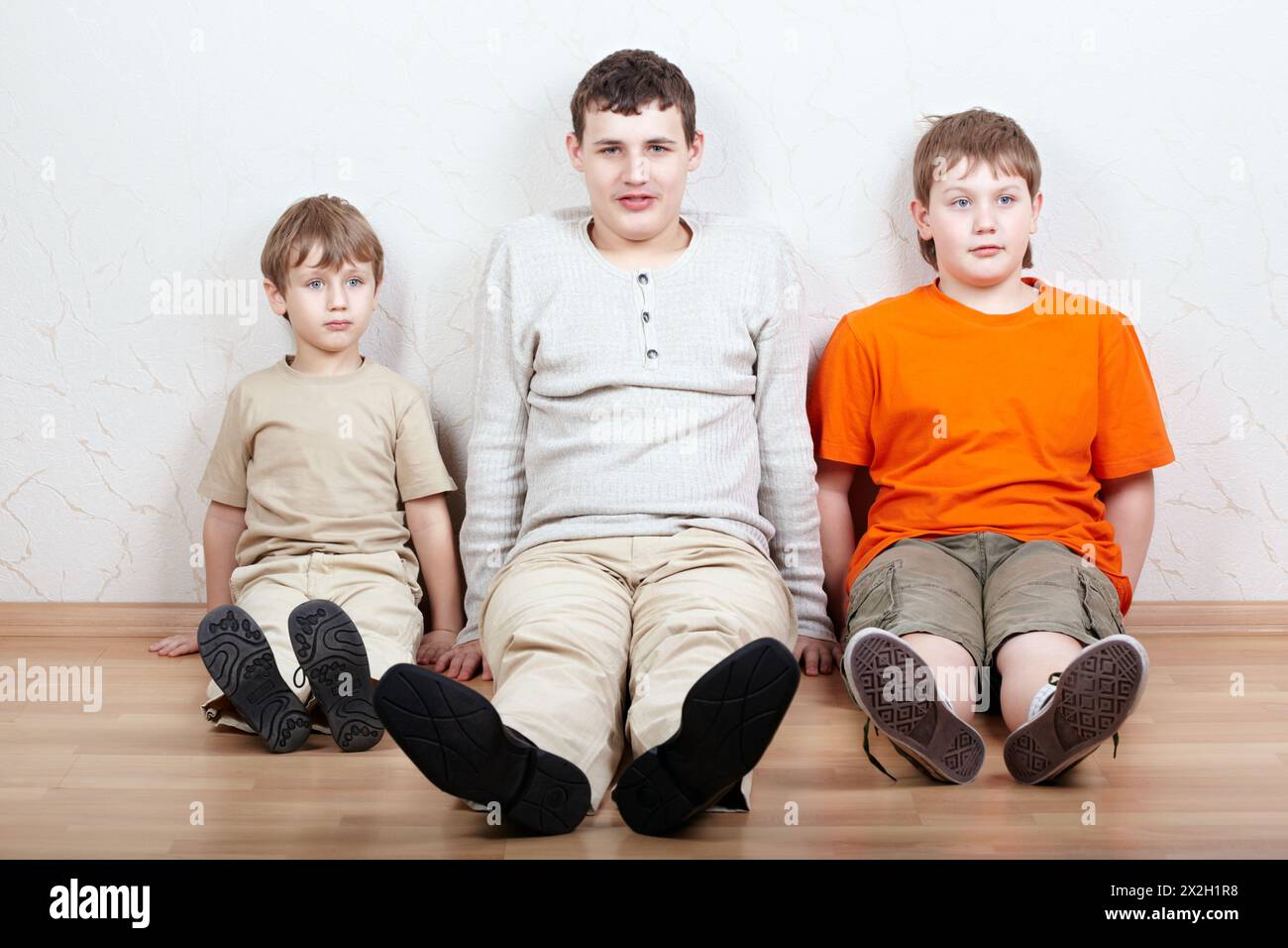 Three boys sit side by side on the floor, legs stretched out before him ...