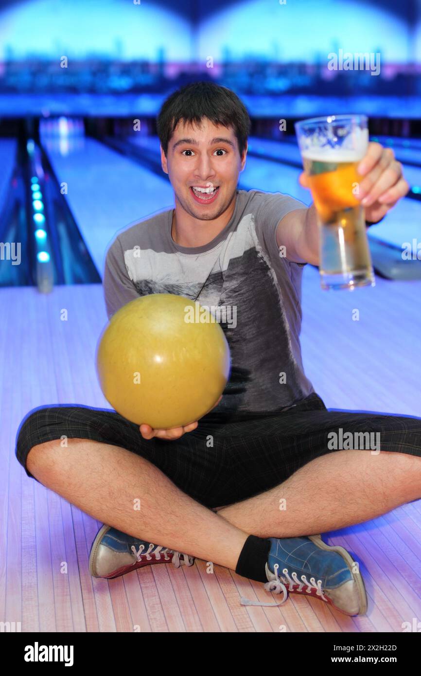 Happy young man sits on floor and holds ball and glass of beer in ...
