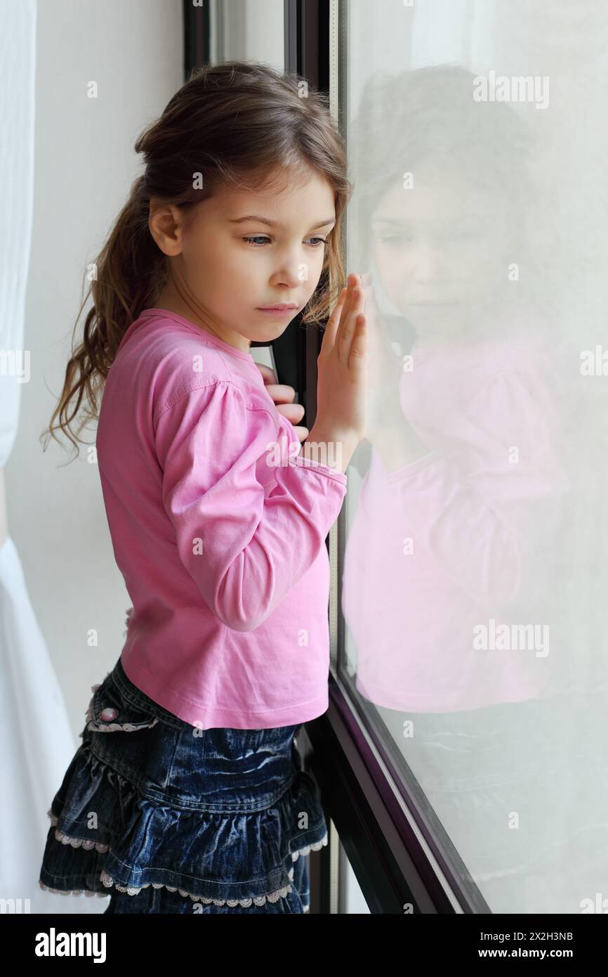 Little beautiful sad girl looks out window and touches glass by hand ...