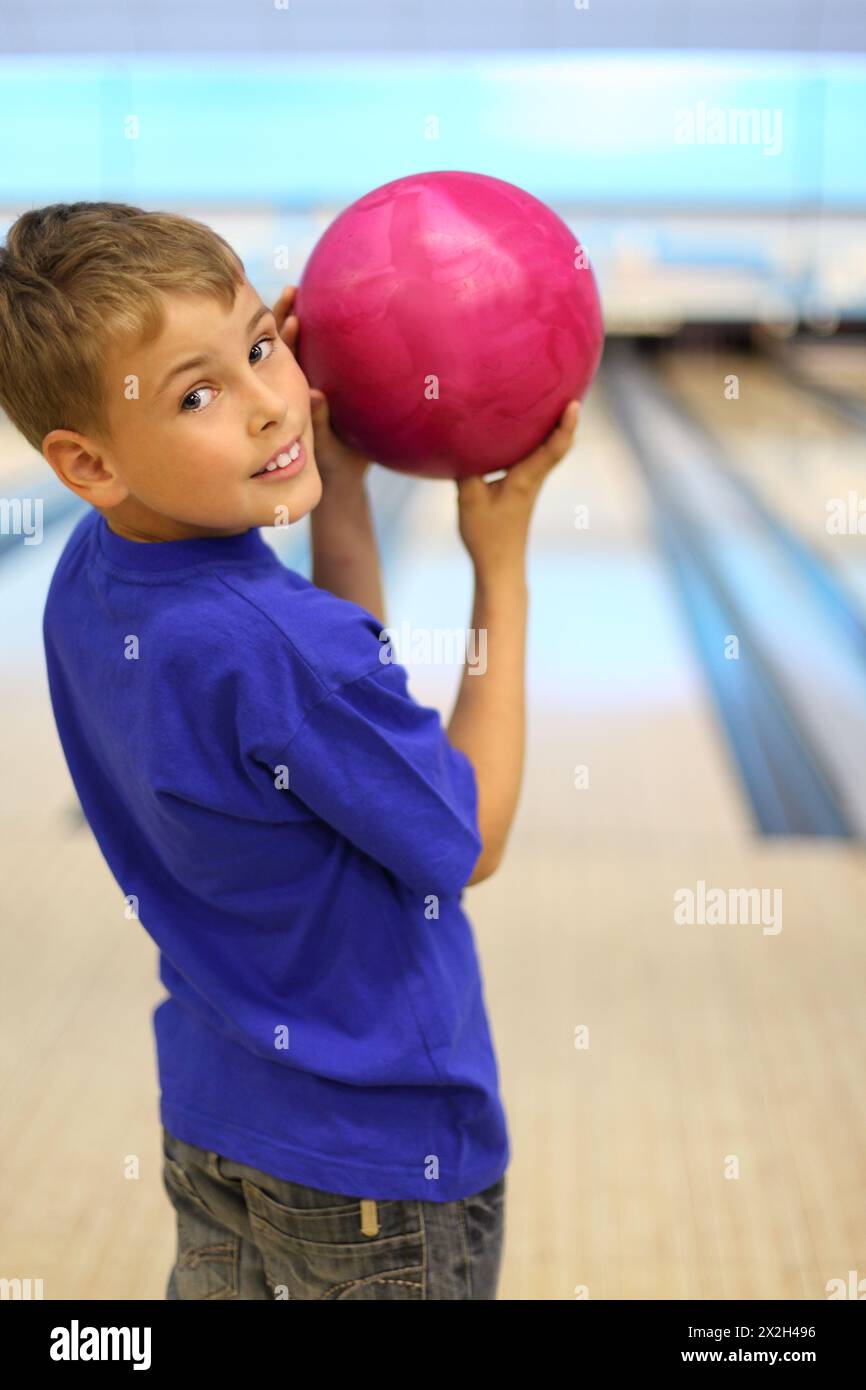 Smiling boy dressed in blue T-shirt holds pink ball in bowling club ...
