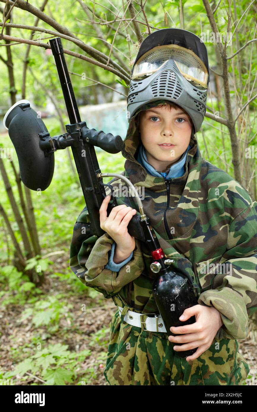 Boy in camouflage stands against brushes on the paintball area raising ...