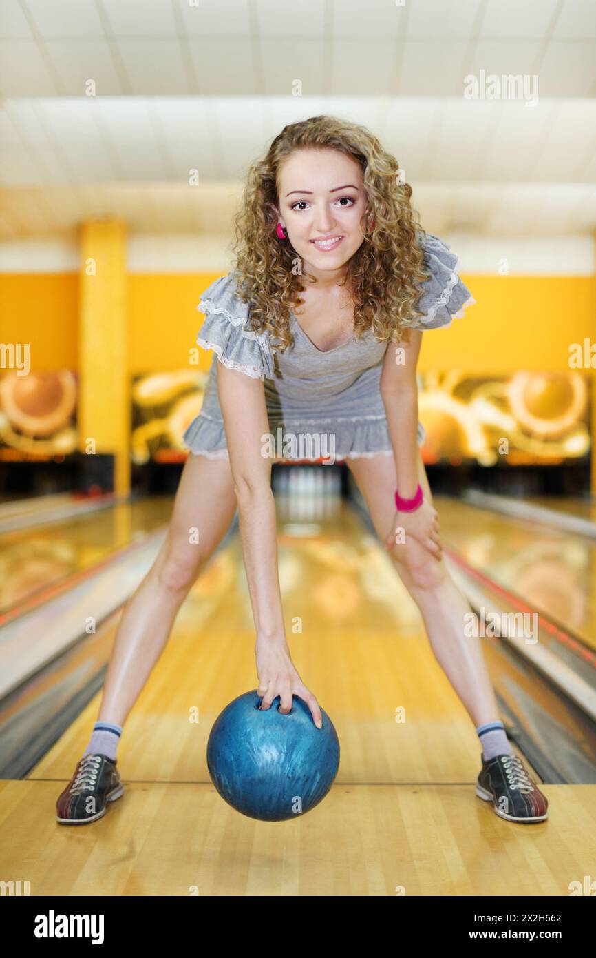 Young beautiful girl holds blue ball and stands bending in bowling club ...