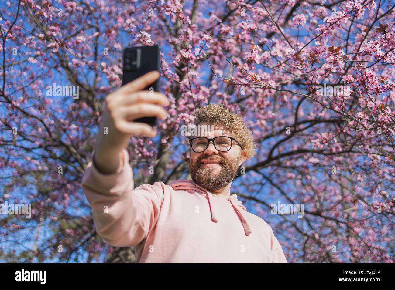 Happy curly man takes selfie against backdrop of flowering tree in ...
