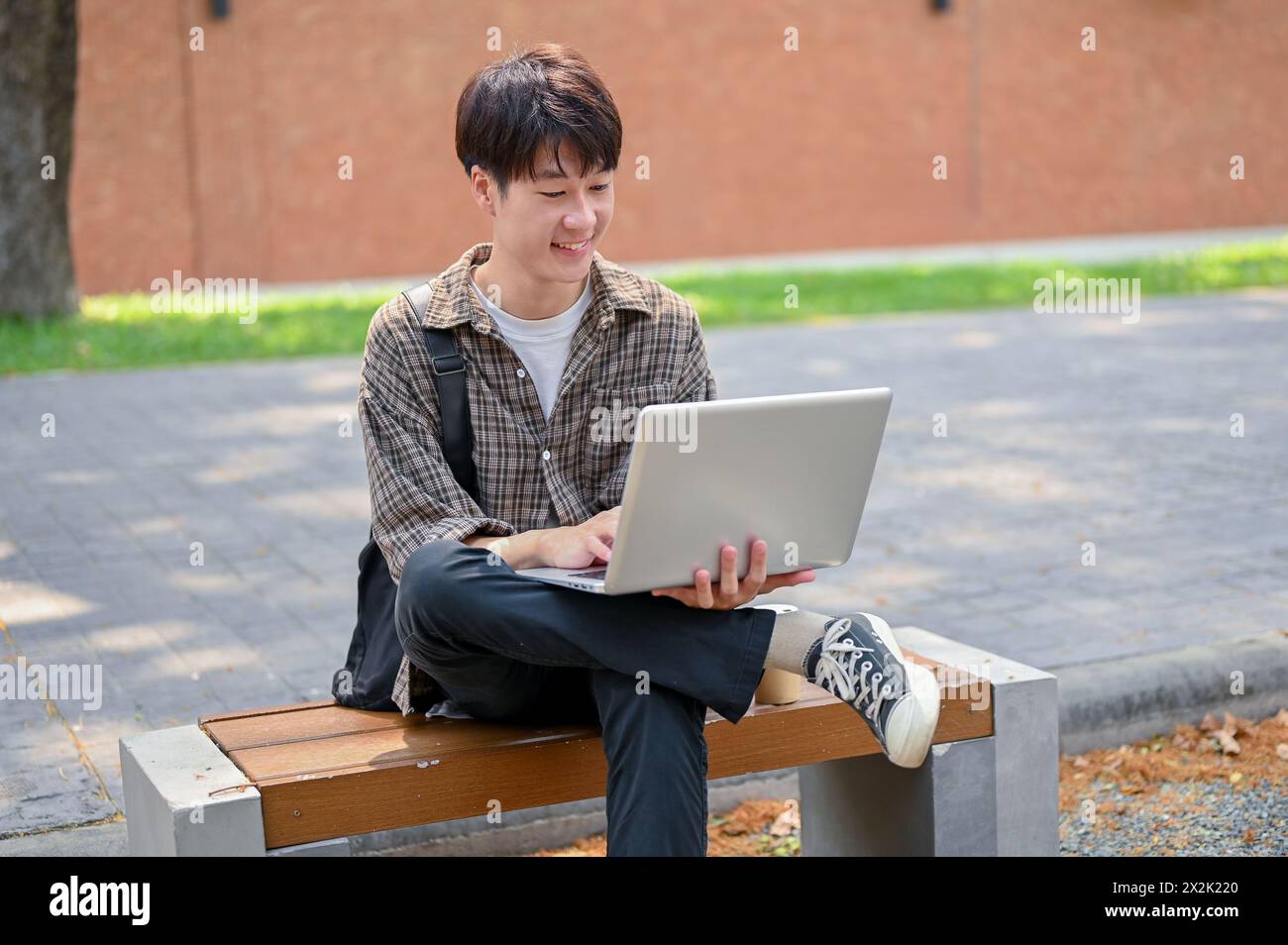 A young Asian male college student using his laptop computer on a bench ...