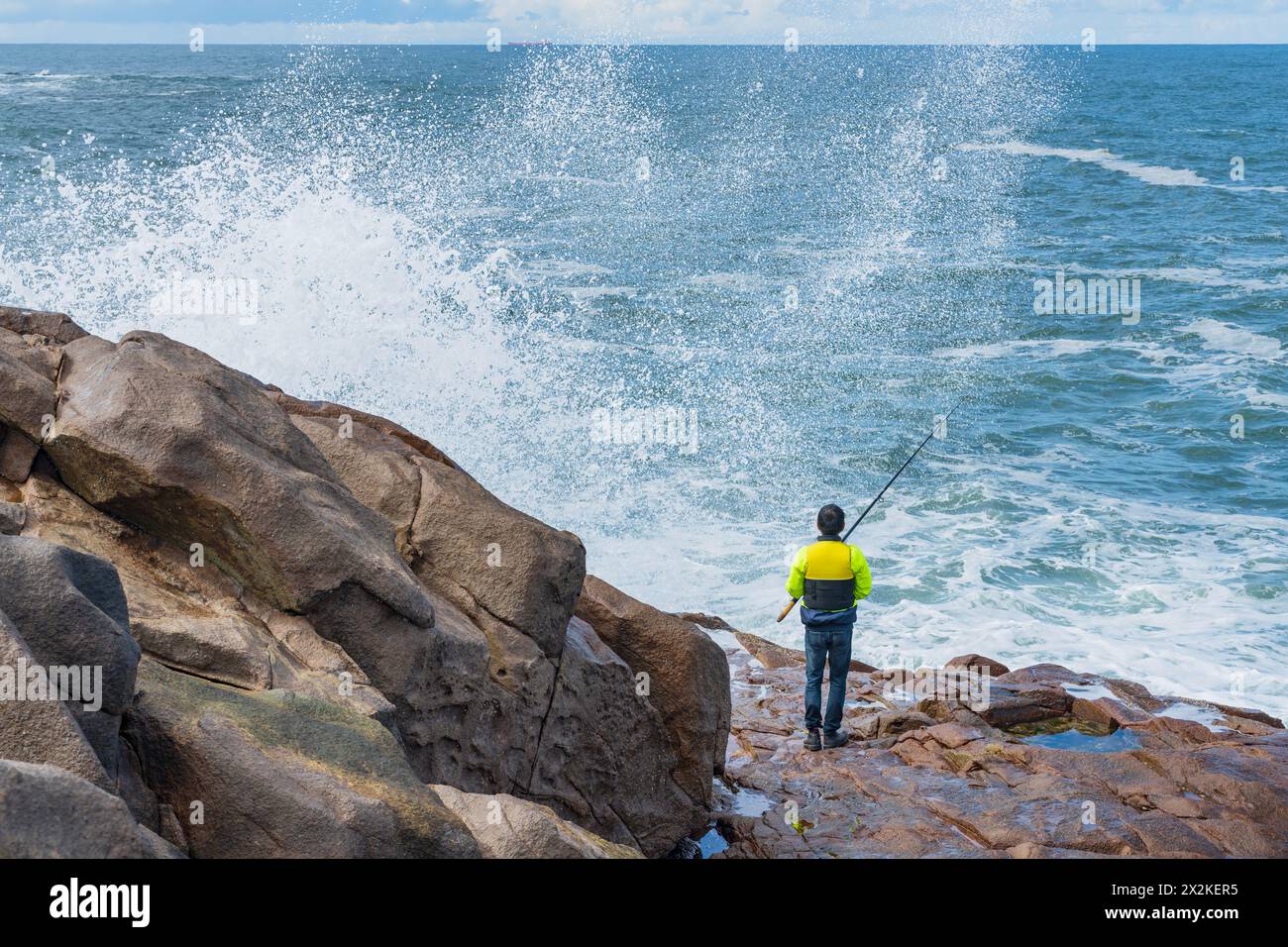 A large wave crashing over a lone rock fisherman standing on a ledge ...