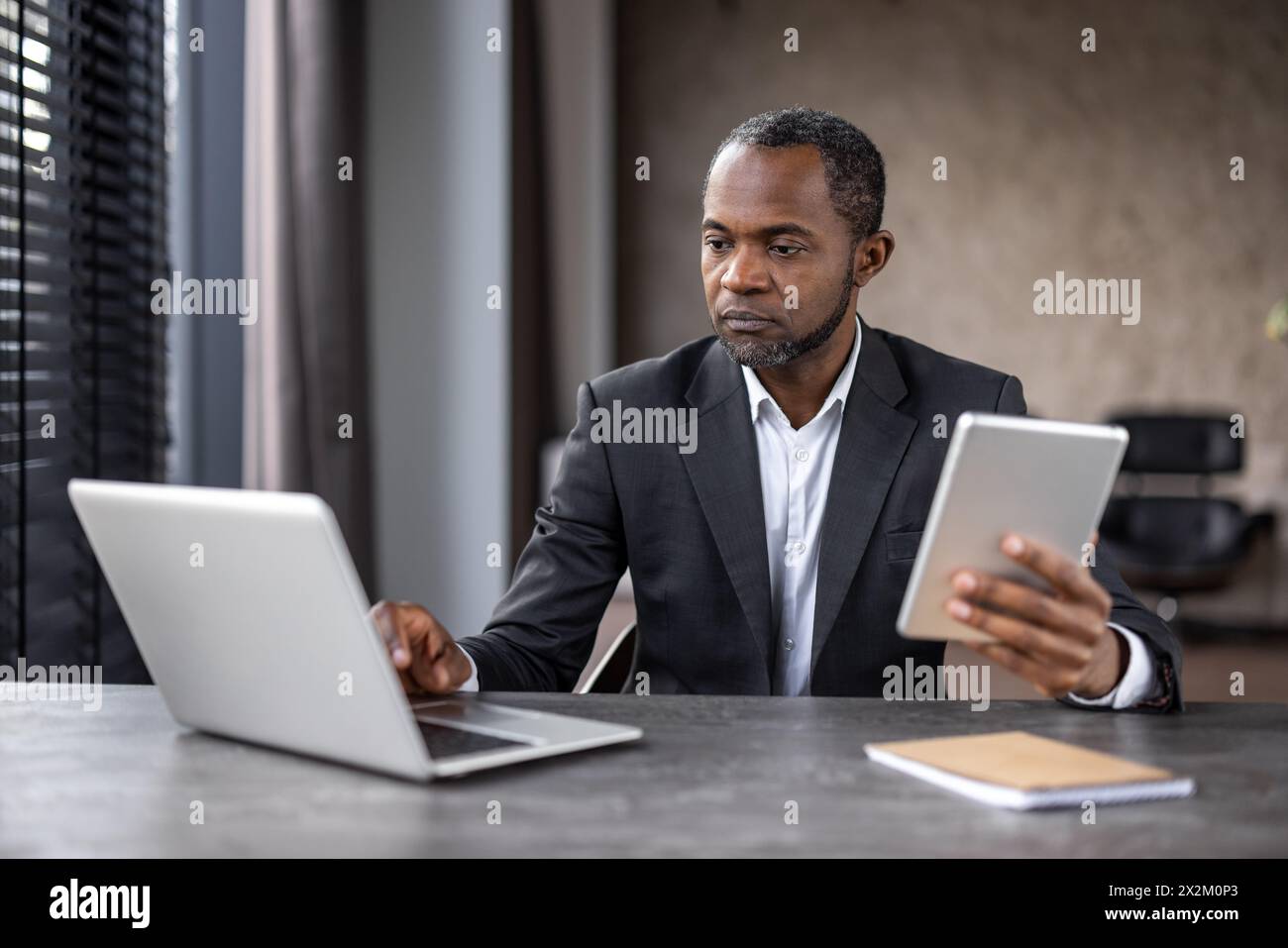 Focused African American businessman uses a laptop and tablet at his ...