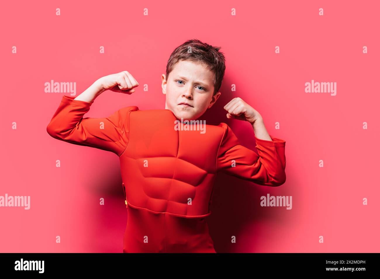 A confident young boy in a red superhero costume flexes his muscles ...