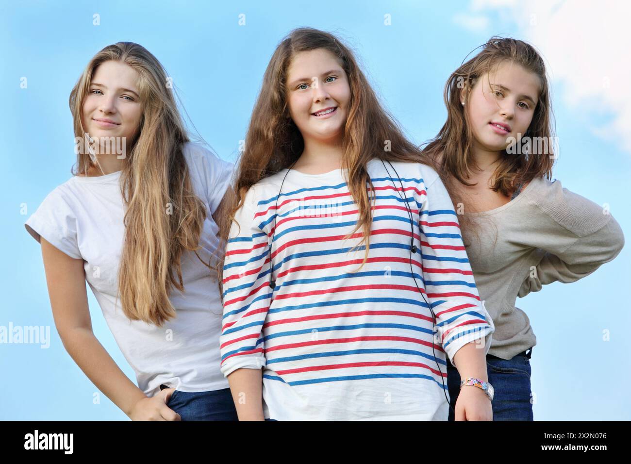 Three happy girls stand together at background of blue sky with clouds ...