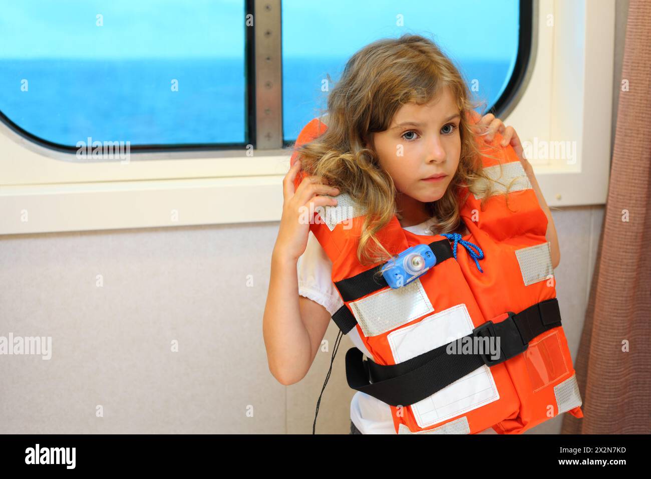 Sad little girl dressed in life jacket stands near window in cabin of ...