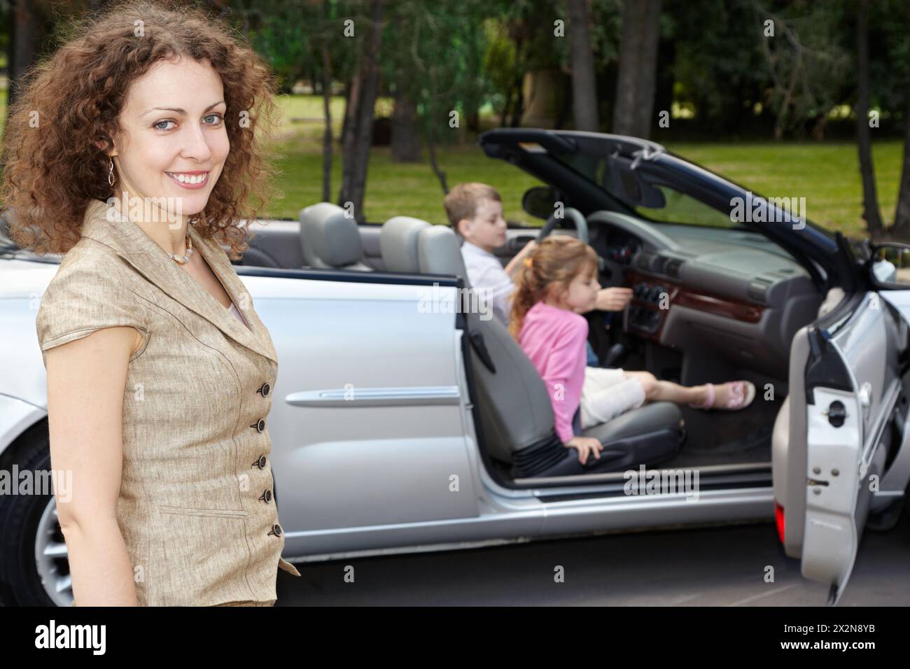 Mother stands half-turned near an open top car, children play inside ...