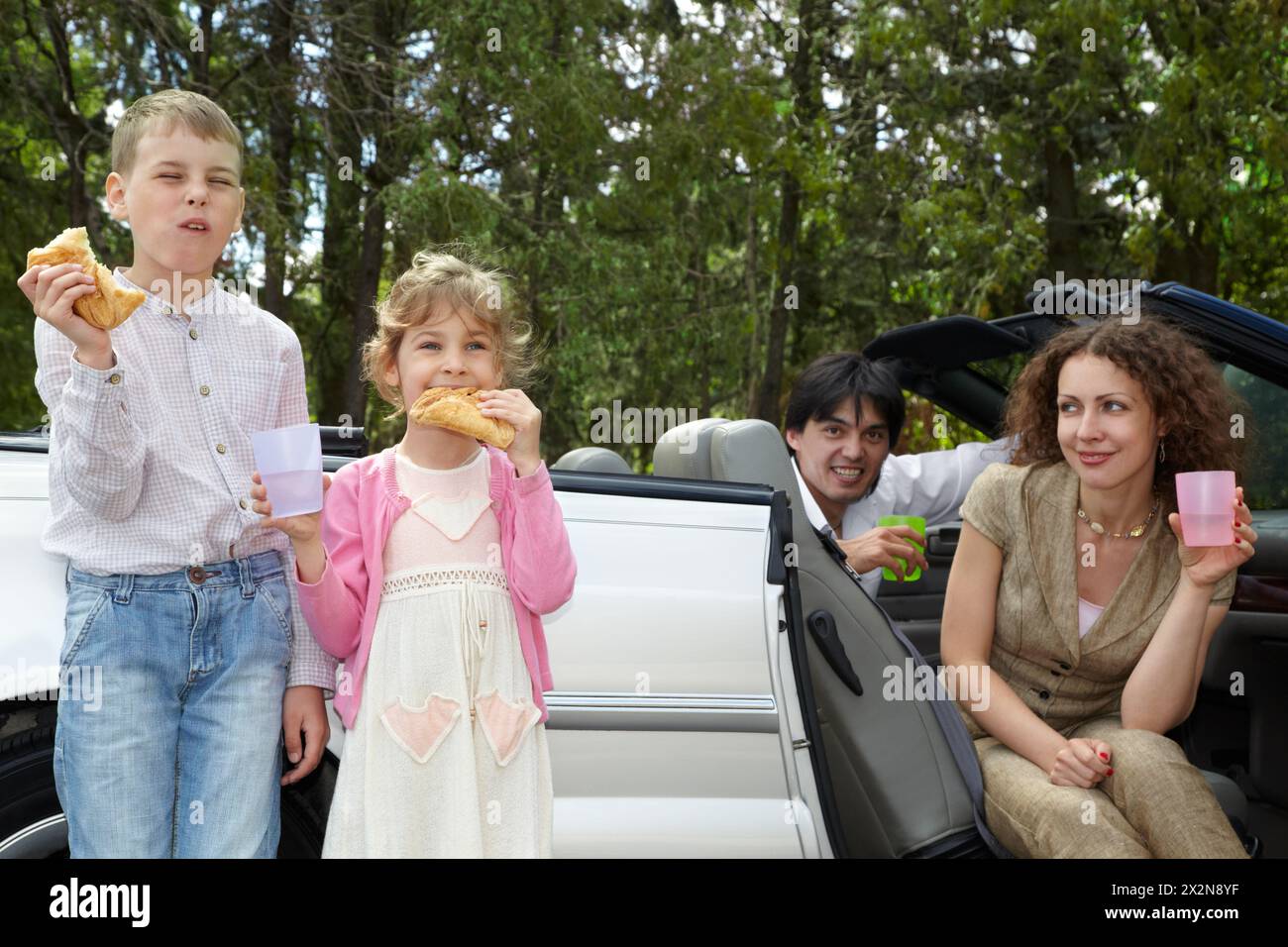 Children stand near the open top car and eat samosa, father and mother ...
