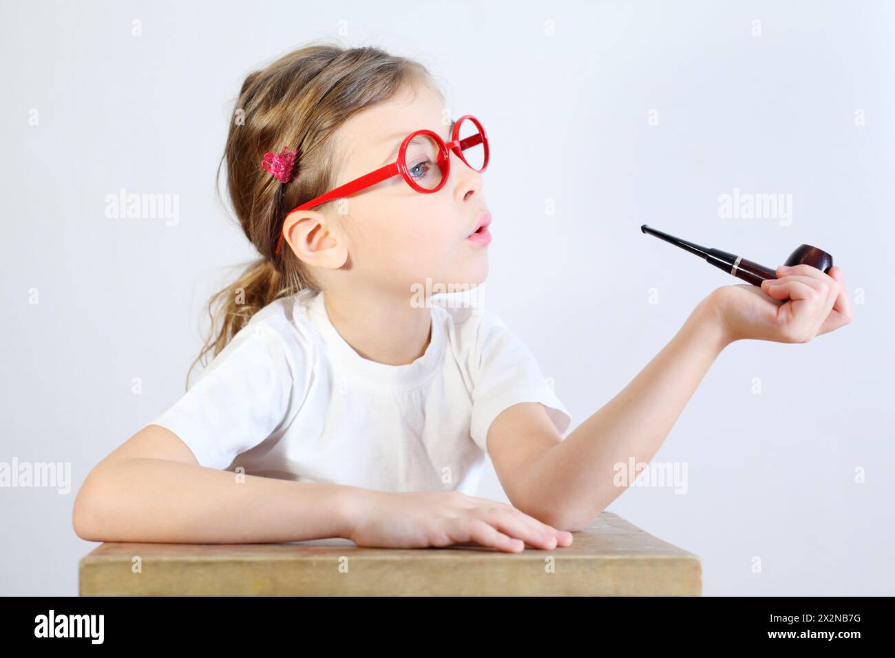 Little girl in big red glasses sits at table and holds pipeful Stock ...