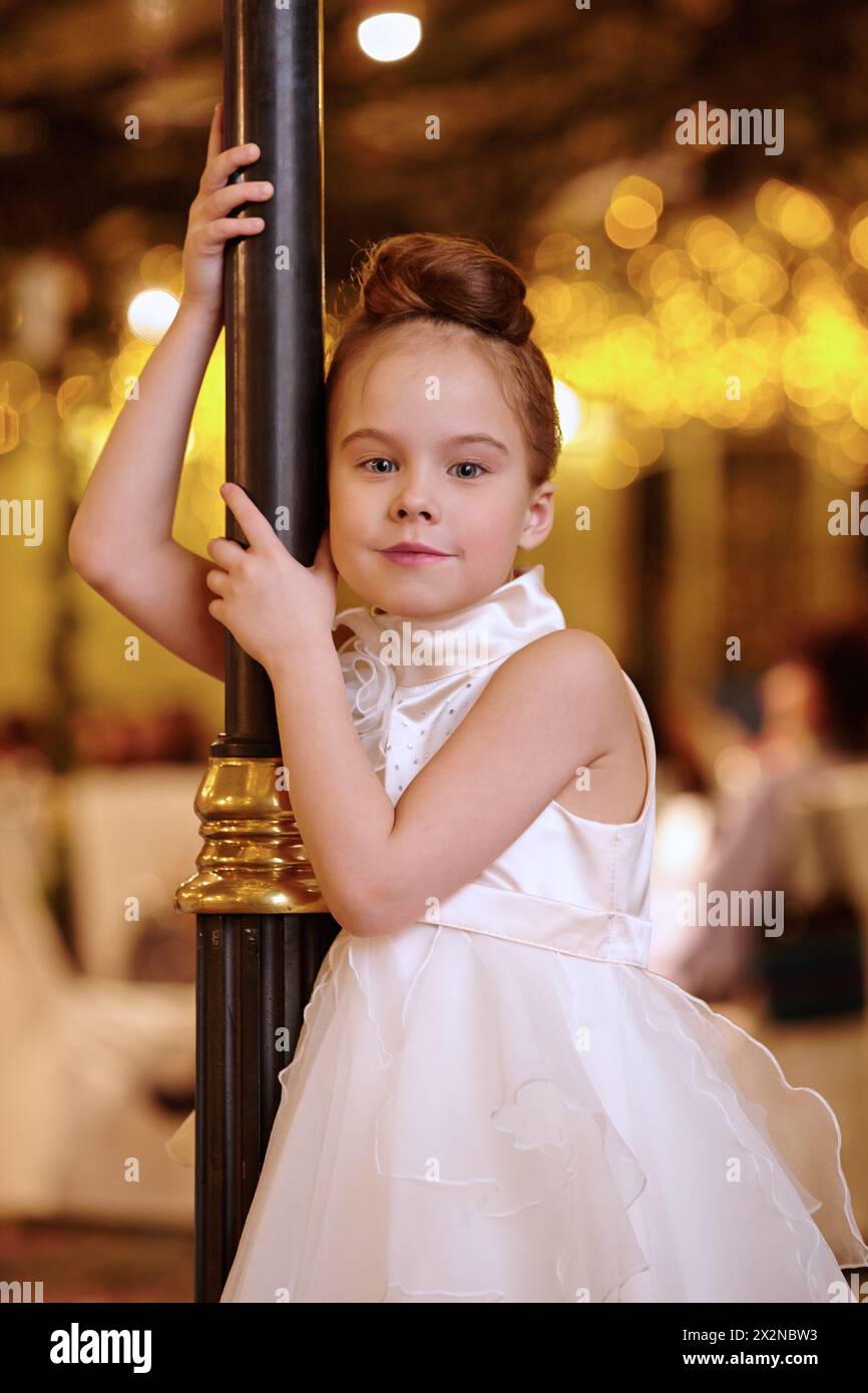 Little girl in white evening dress stands leaning to decorative ...