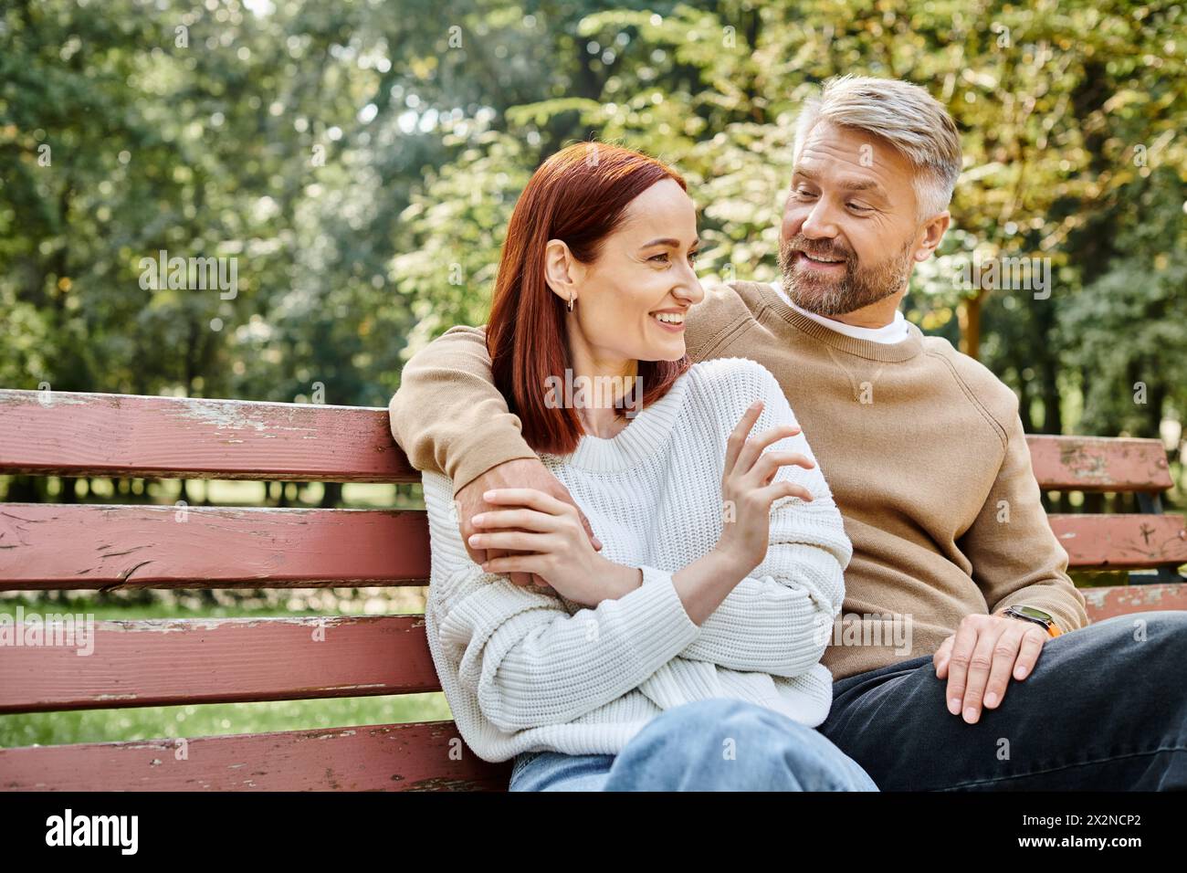 A loving adult couple in casual attire sitting together on a park bench ...