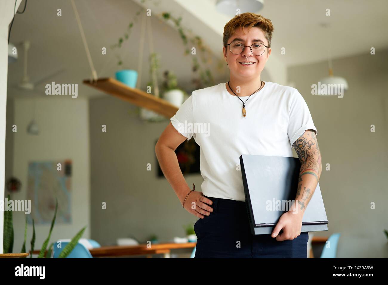 Transgender professional in bright office smiles, holding laptop, inked ...