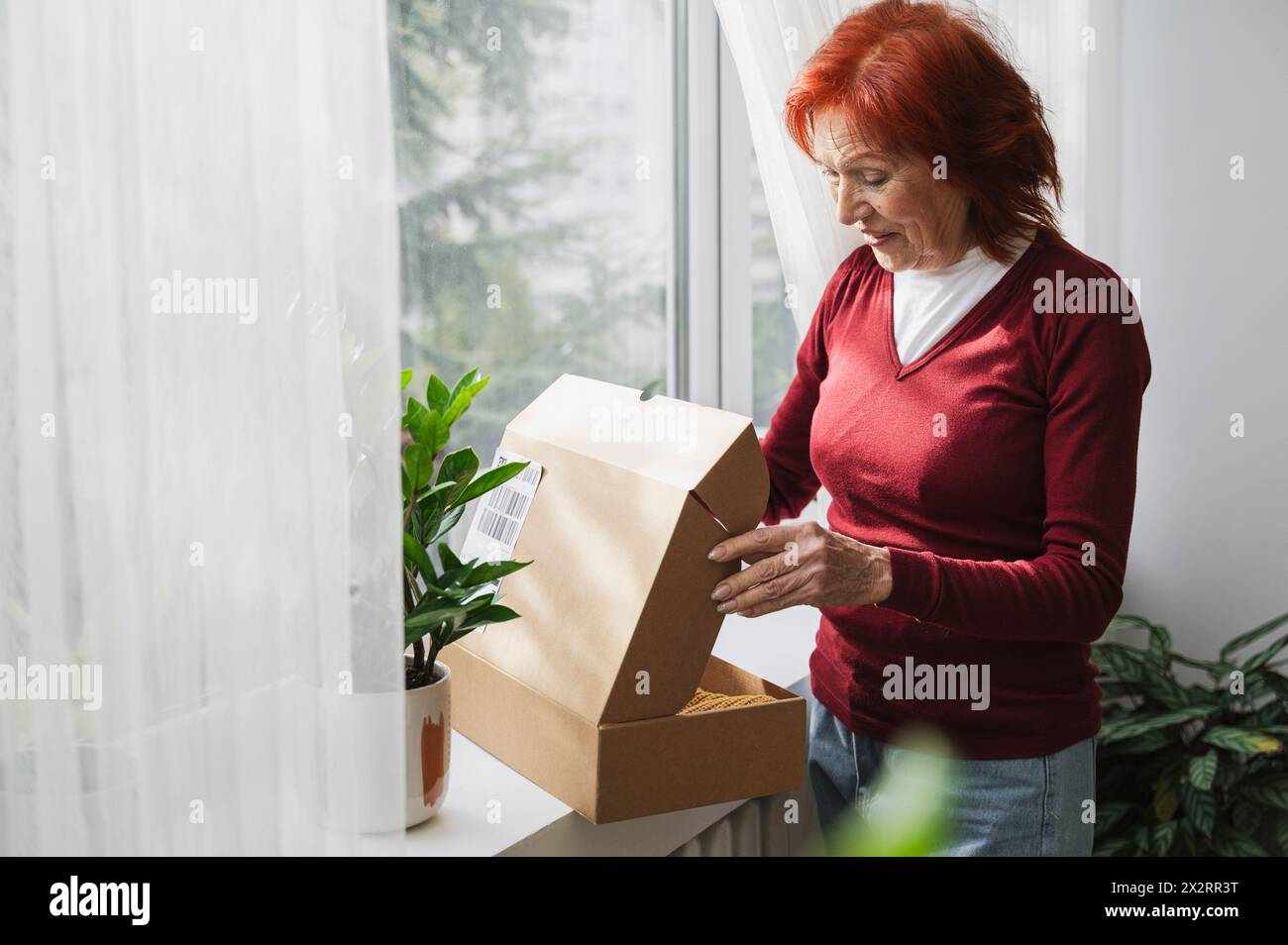 Excited senior woman opening cardboard box standing by window at home ...