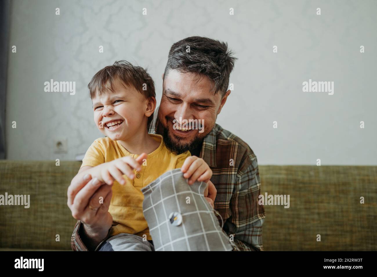 Happy father teaching son to sew button on gray colored cloth at home ...