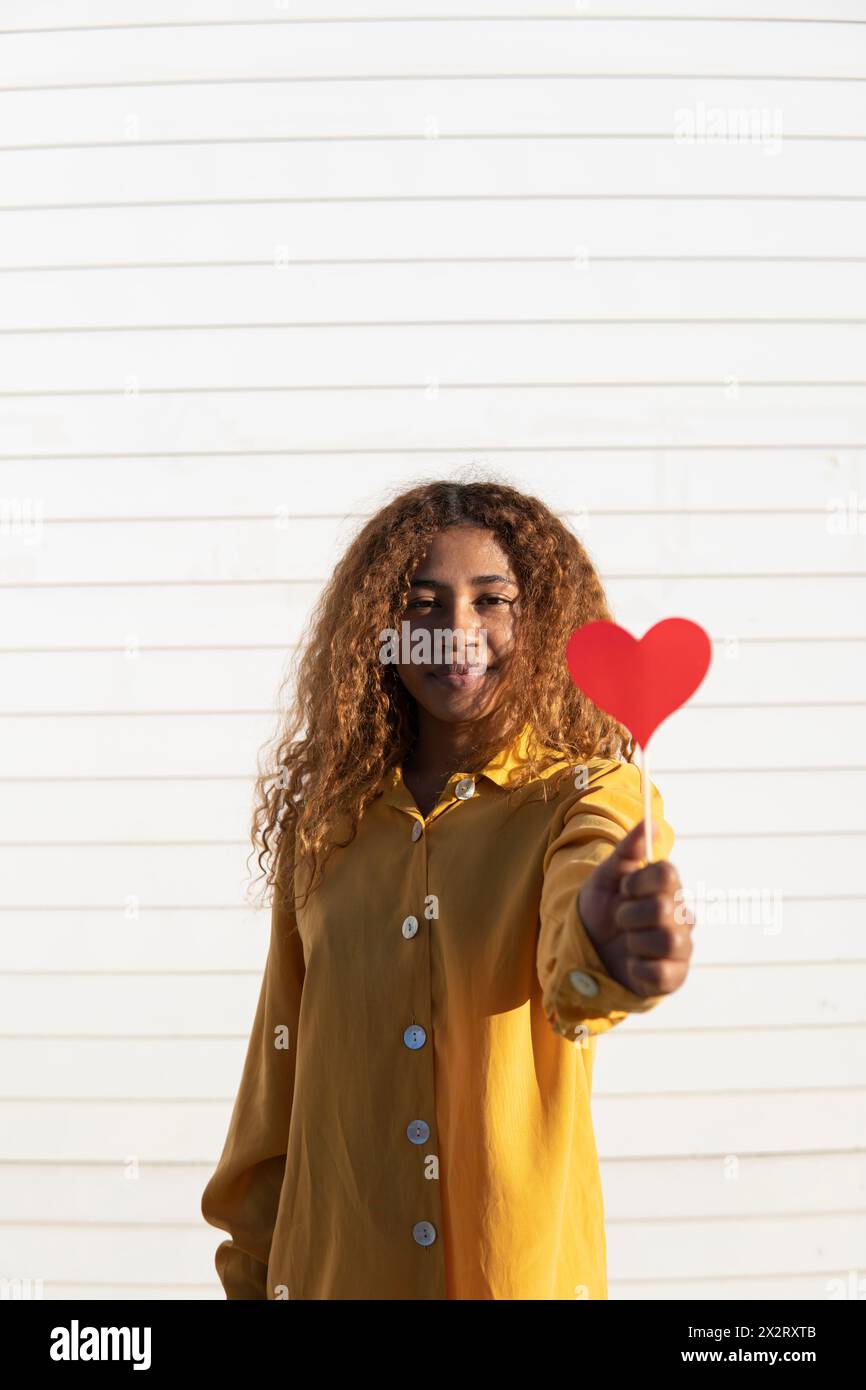 Young curly haired woman giving red heart shape prop in front of white ...