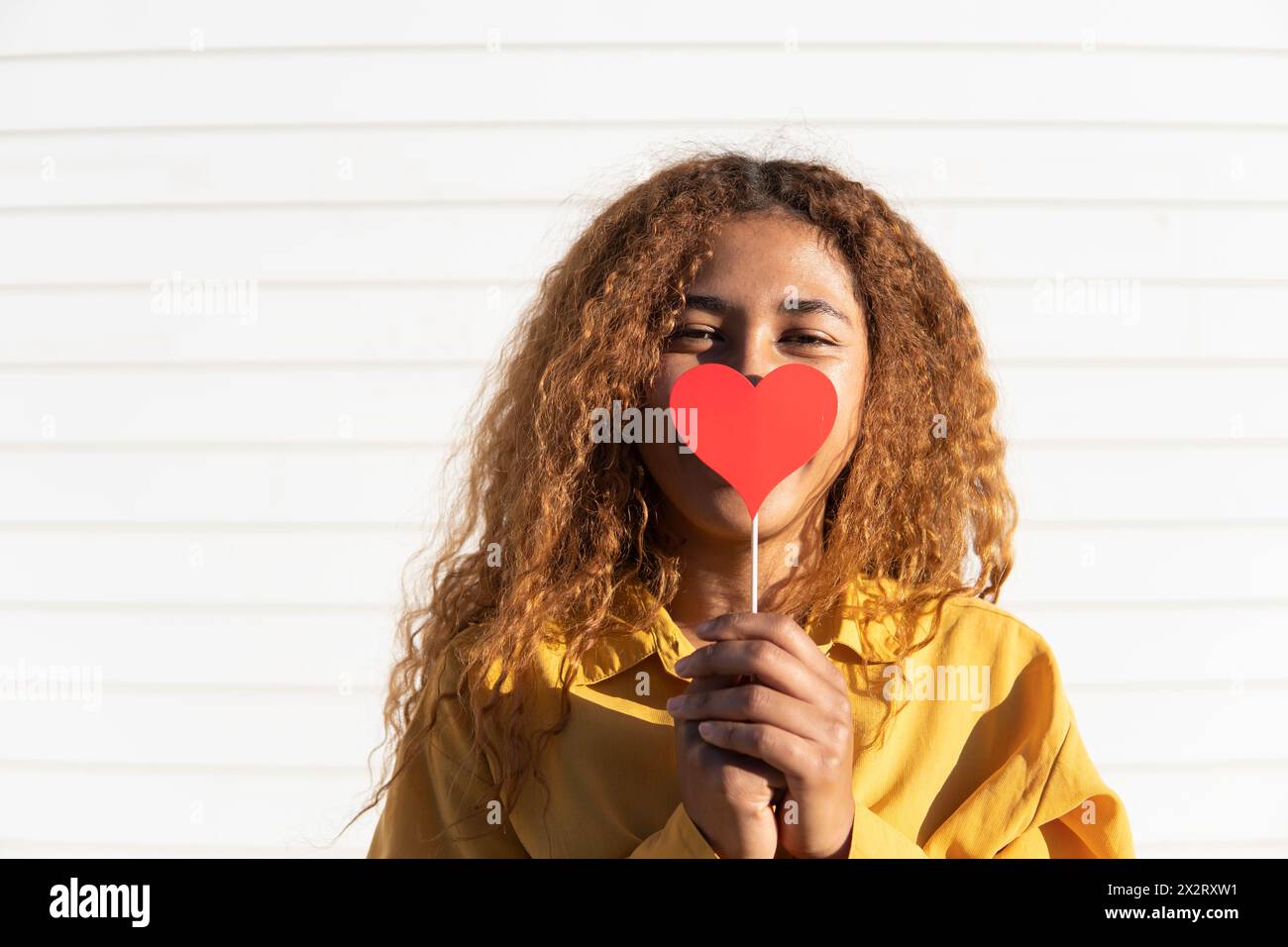 Young woman holding red heart shape prop over mouth in front of white ...