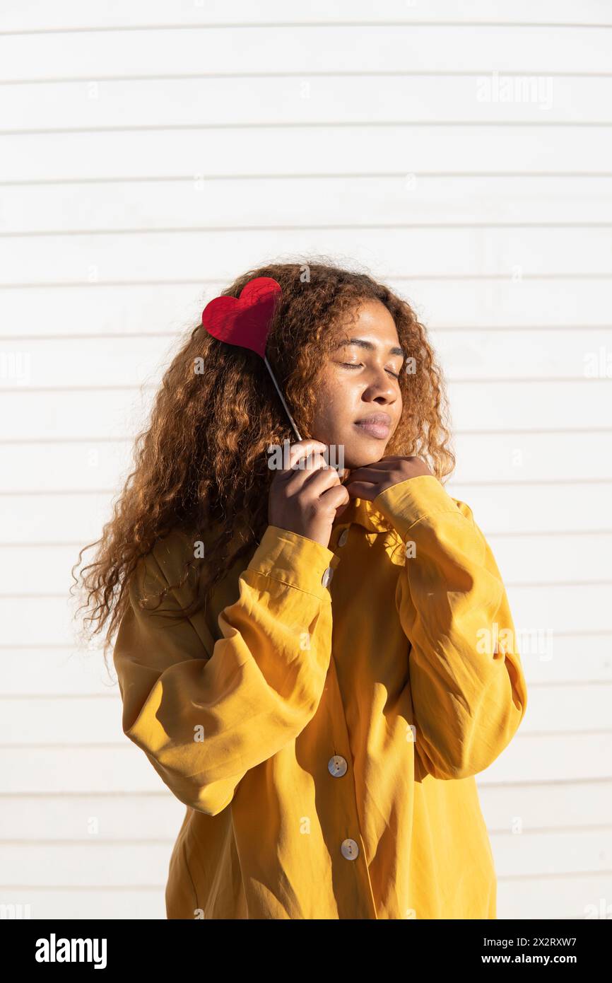Young curly haired woman with eyes closed holding red heart shape prop ...
