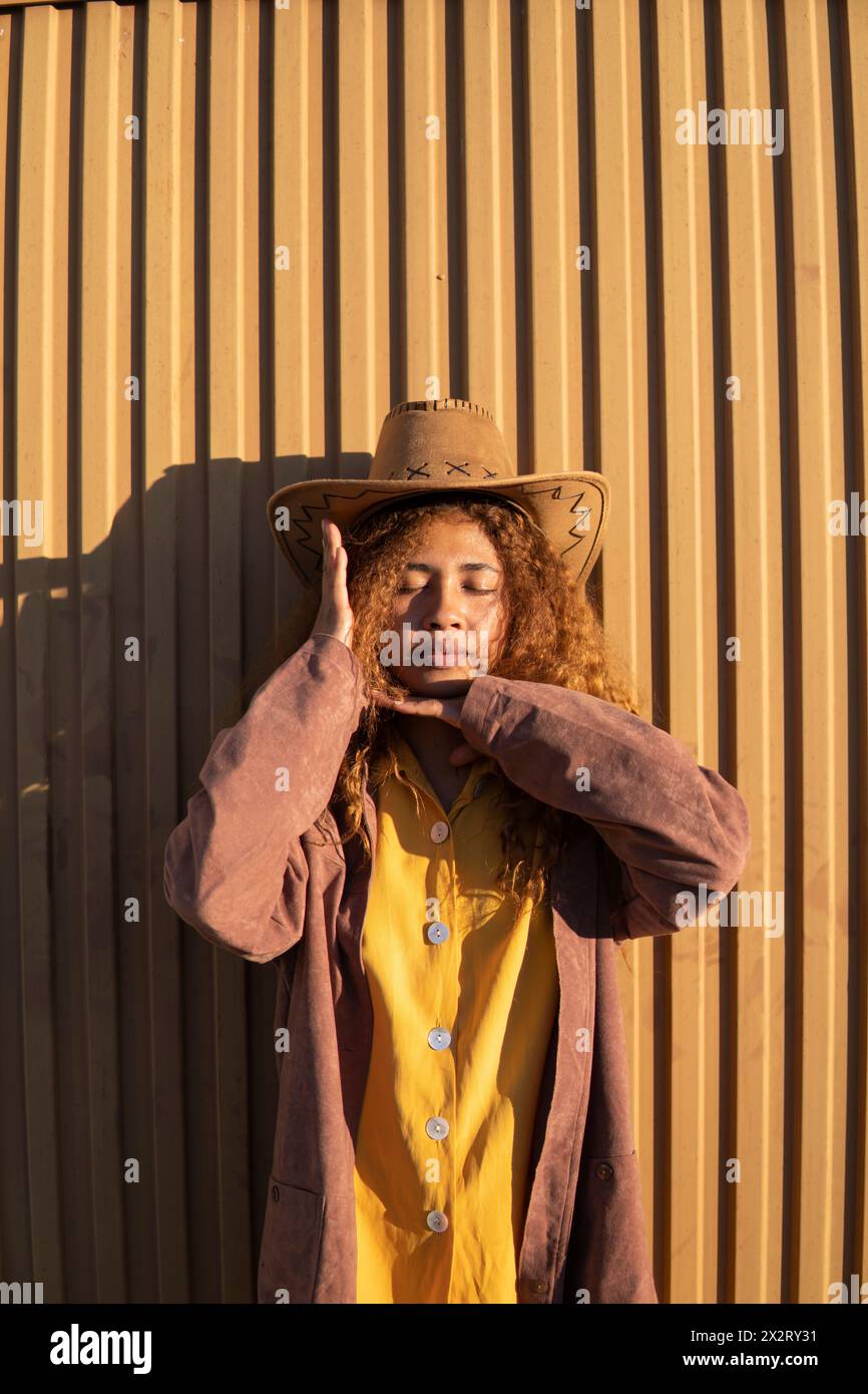 Woman with eyes closed wearing jacket and cowboy hat standing in front of orange metal wall ...