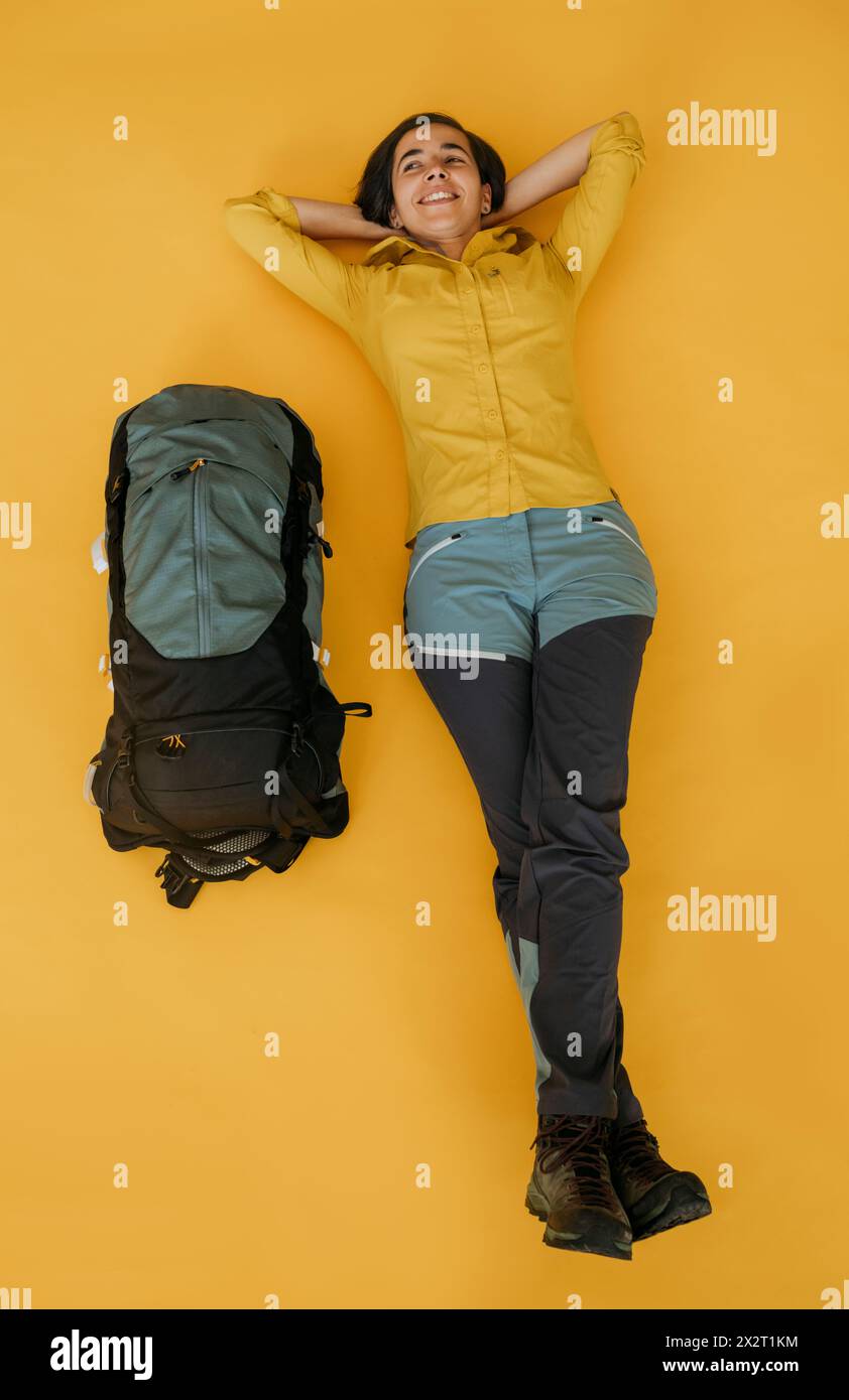 Thoughtful woman lying down near backpack on yellow background Stock ...