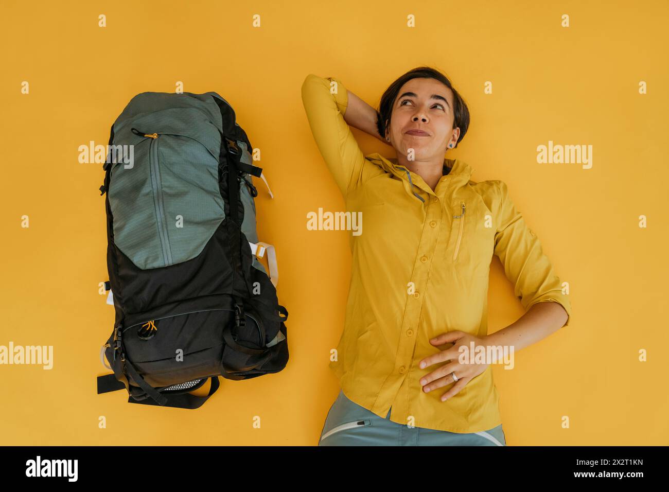 Thoughtful woman lying with backpack on yellow background Stock Photo ...