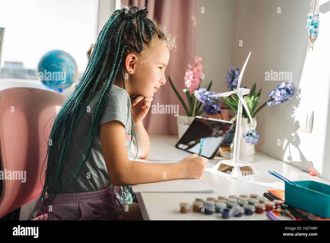 Girl with dyed long braided hair writing in book by wind turbine model ...