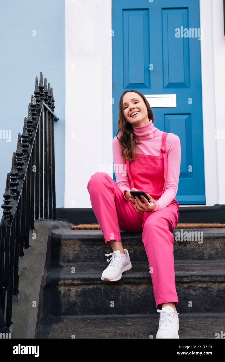 Happy woman wearing pink bib overalls with smart phone sitting on steps ...