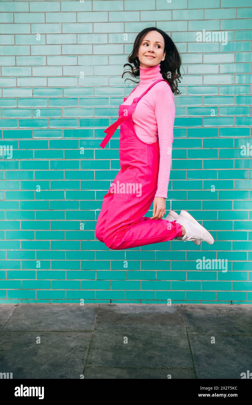 Smiling woman wearing pink bib overalls jumping in front of blue brick ...