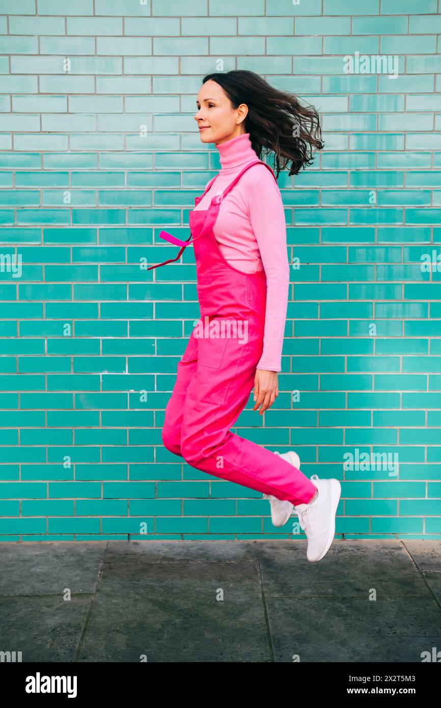 Woman wearing pink bib overalls jumping in front of blue brick wall ...